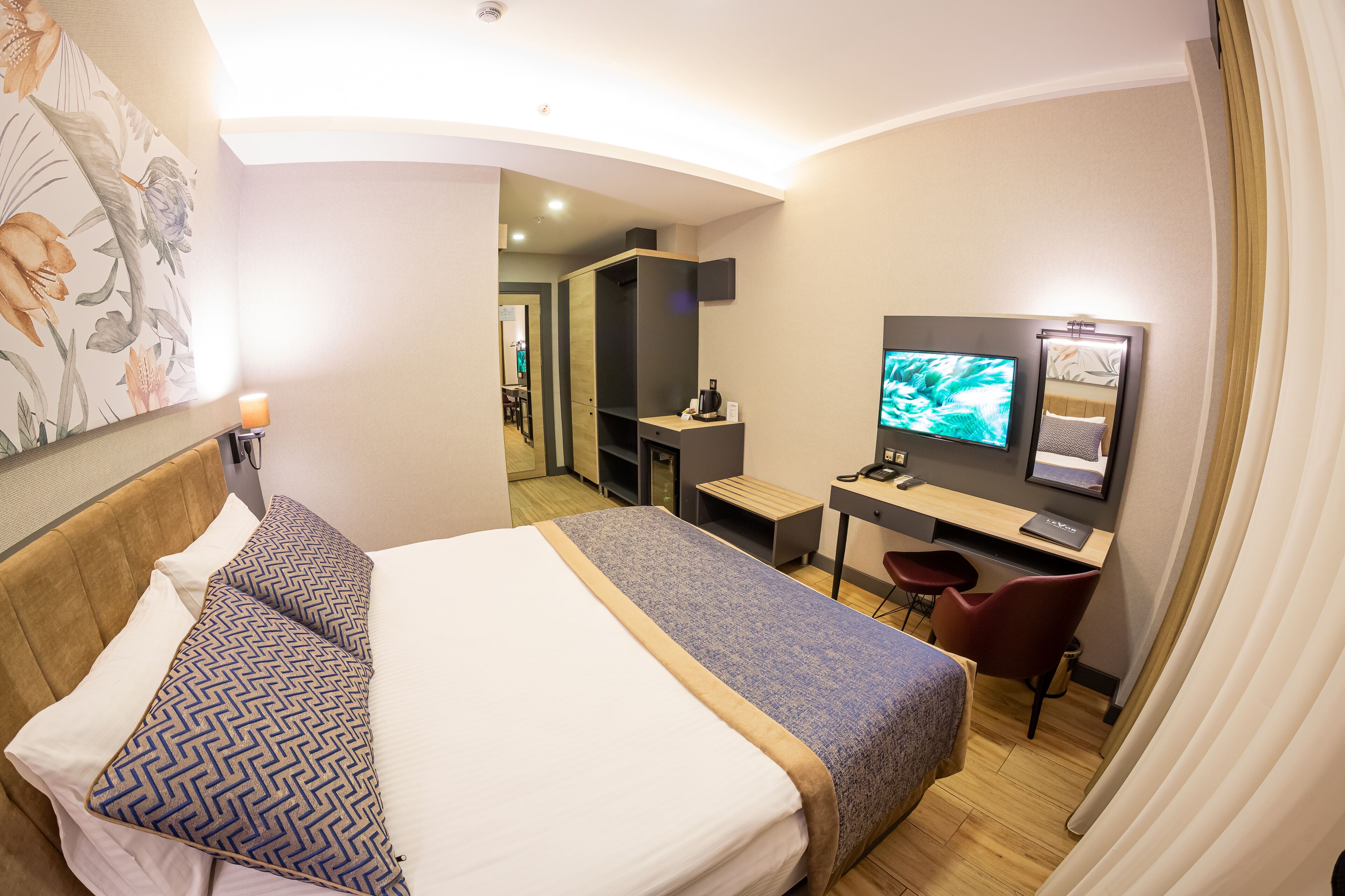 Quarto de hotel com cama, escrivaninha, cadeira, televisão e banheiro ao fundo.