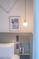 Quarto de hotel com uma cama, mesa de cabeceira com livros, luminária na parede e um quadro emoldurado.
