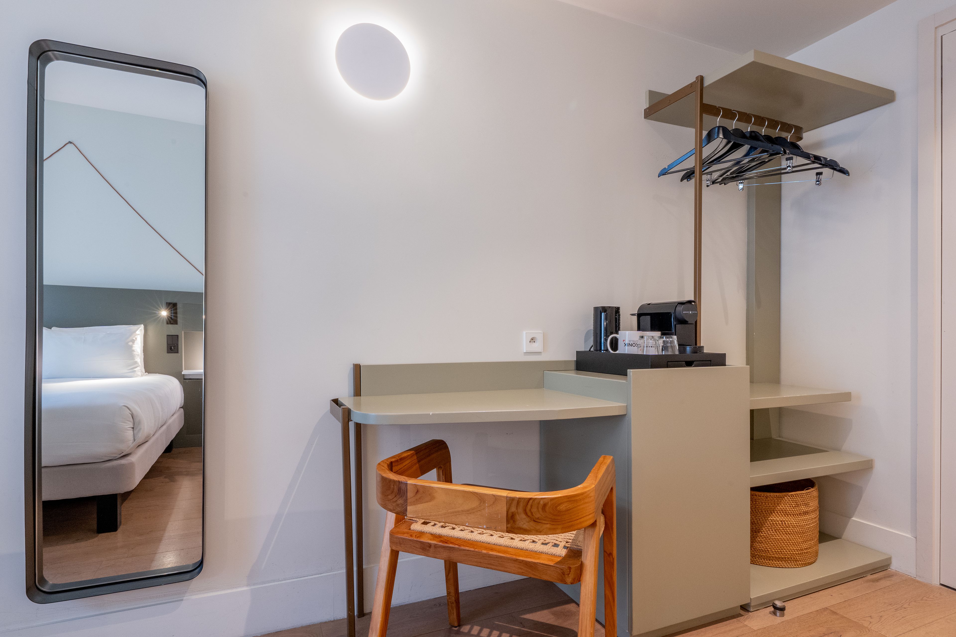 Quarto de hotel com cama, escrivaninha, cadeira, espelho e prateleira com roupas e cafeteira.