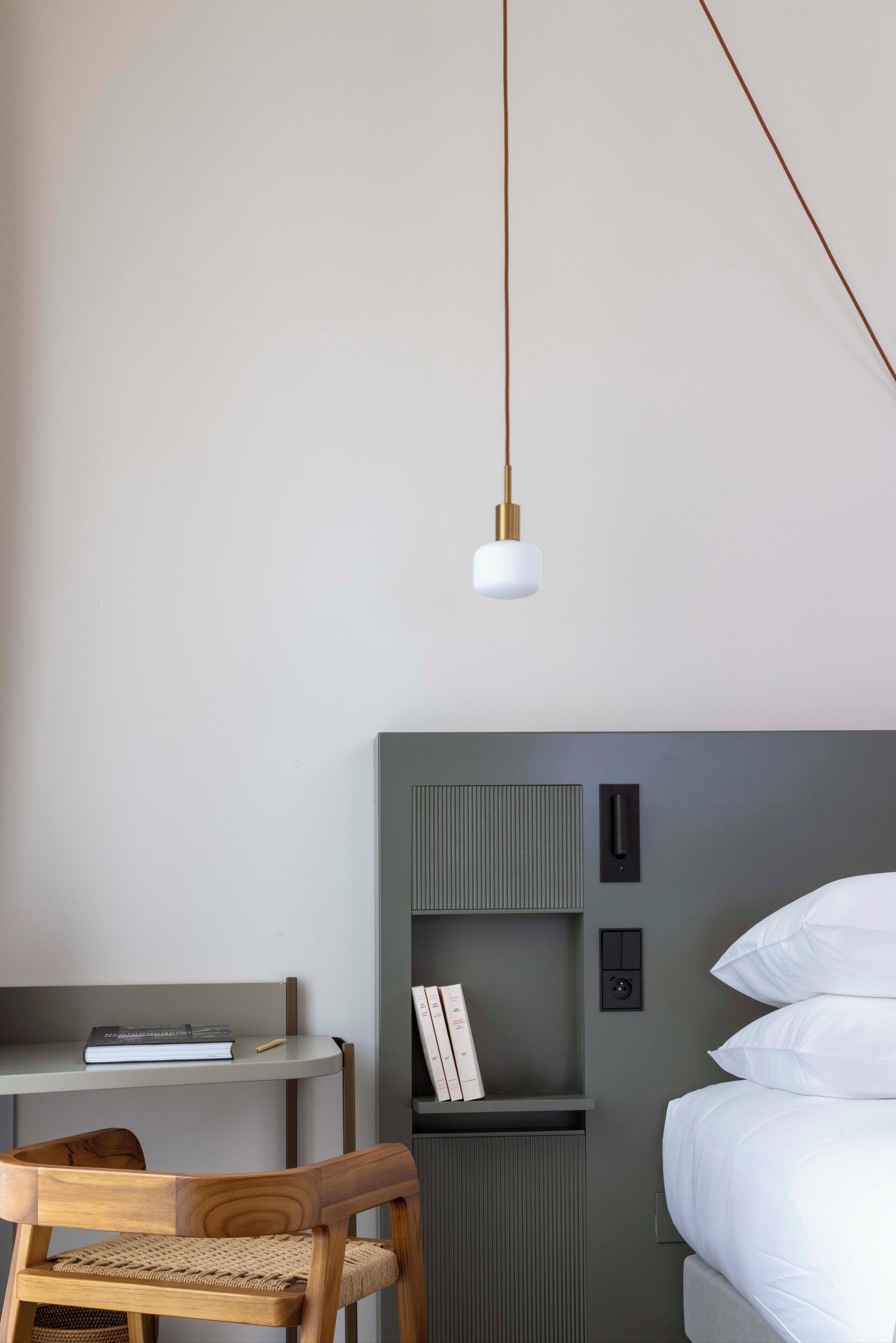 Quarto de hotel moderno com cama, escrivaninha e cadeira, e luminária suspensa.