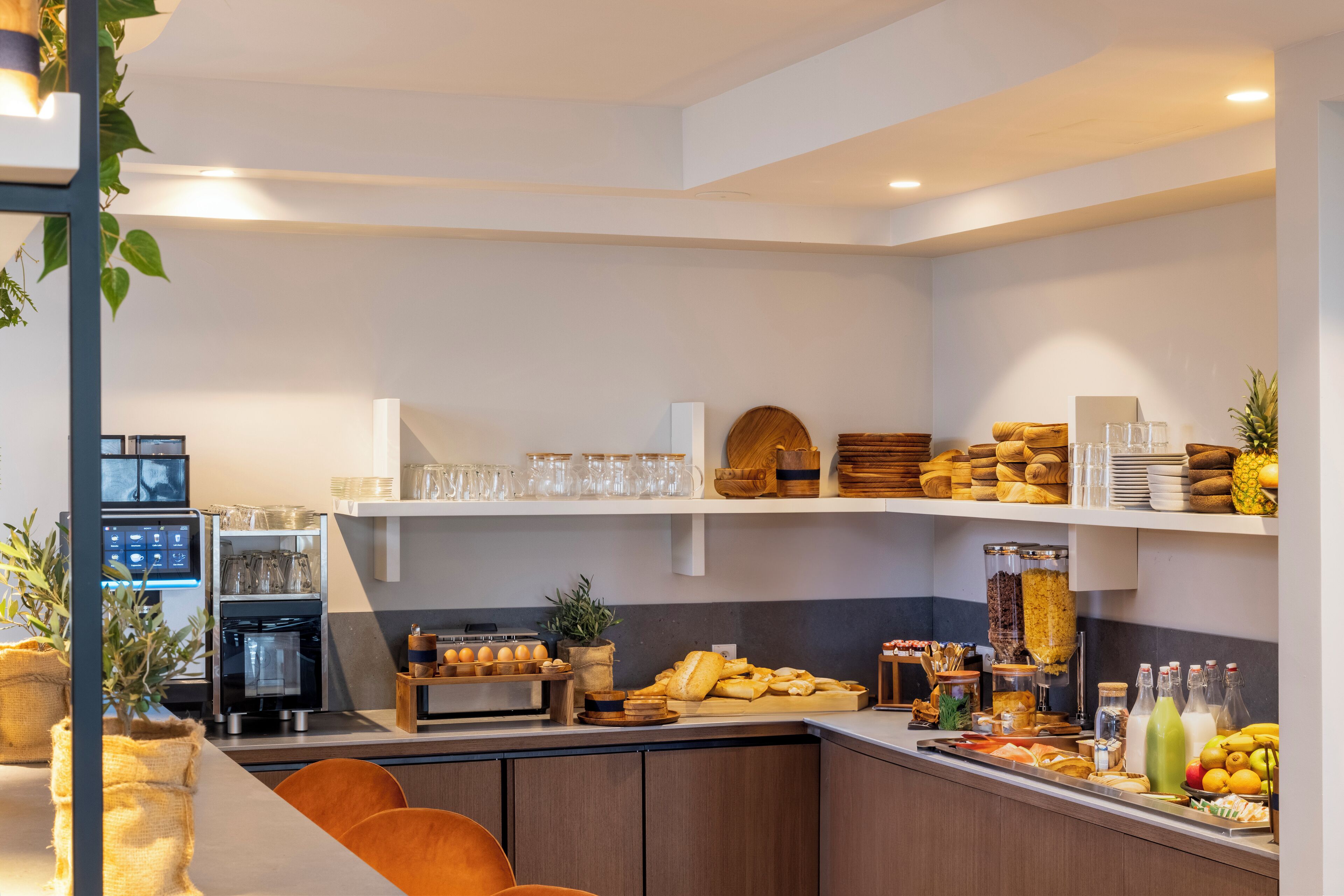 Um buffet de café da manhã com diversas opções de comida e bebidas, uma máquina de café e uma área de estar com cadeiras laranjas.