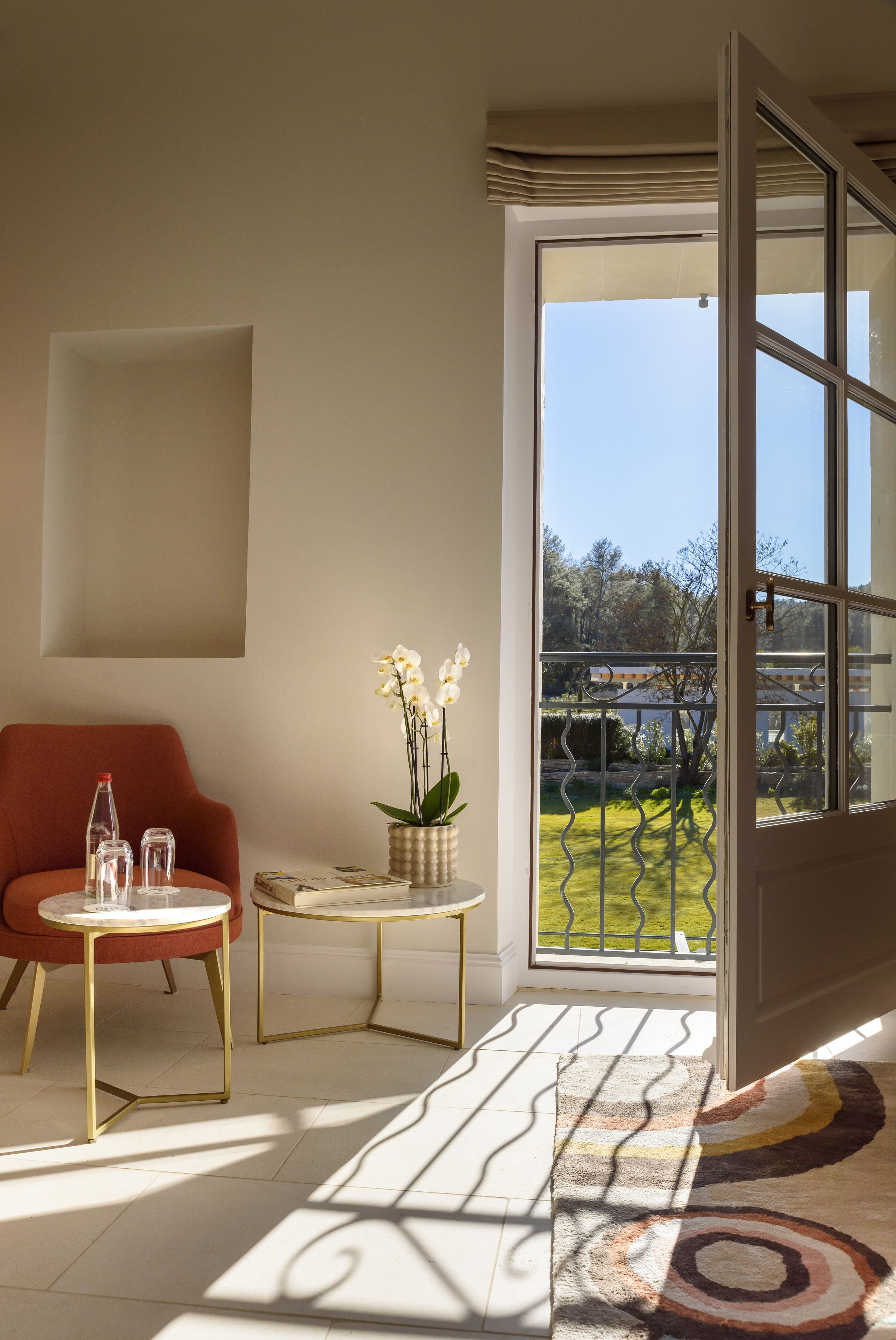 Quarto com uma cadeira vermelha, uma mesa redonda pequena, um vaso com flores brancas e vista para um jardim através de uma porta de vidro.