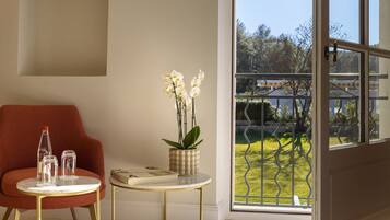 Quarto com uma cadeira vermelha, uma mesa redonda pequena, um vaso com flores brancas e vista para um jardim através de uma porta de vidro.