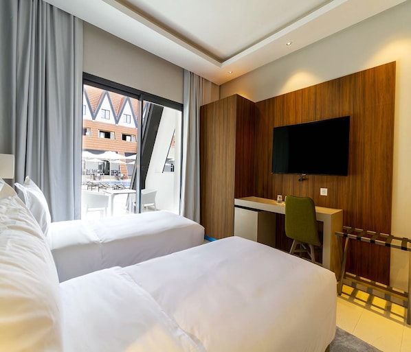 Quarto de hotel com duas camas, uma mesa, uma cadeira, televisão e vista para o exterior.