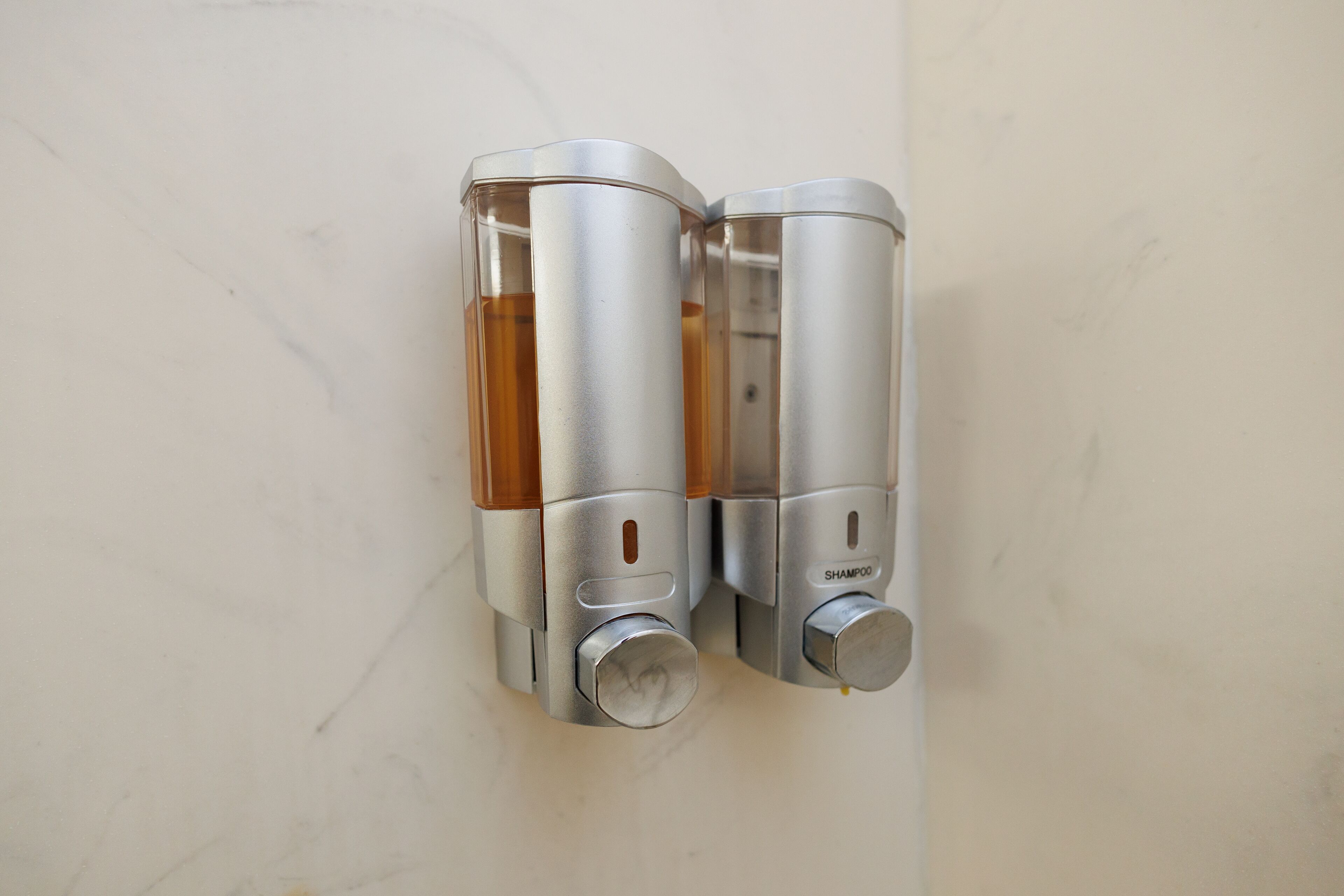 Dois dispensadores de champô fixados na parede, com acabamento metálico e líquido castanho no interior.
