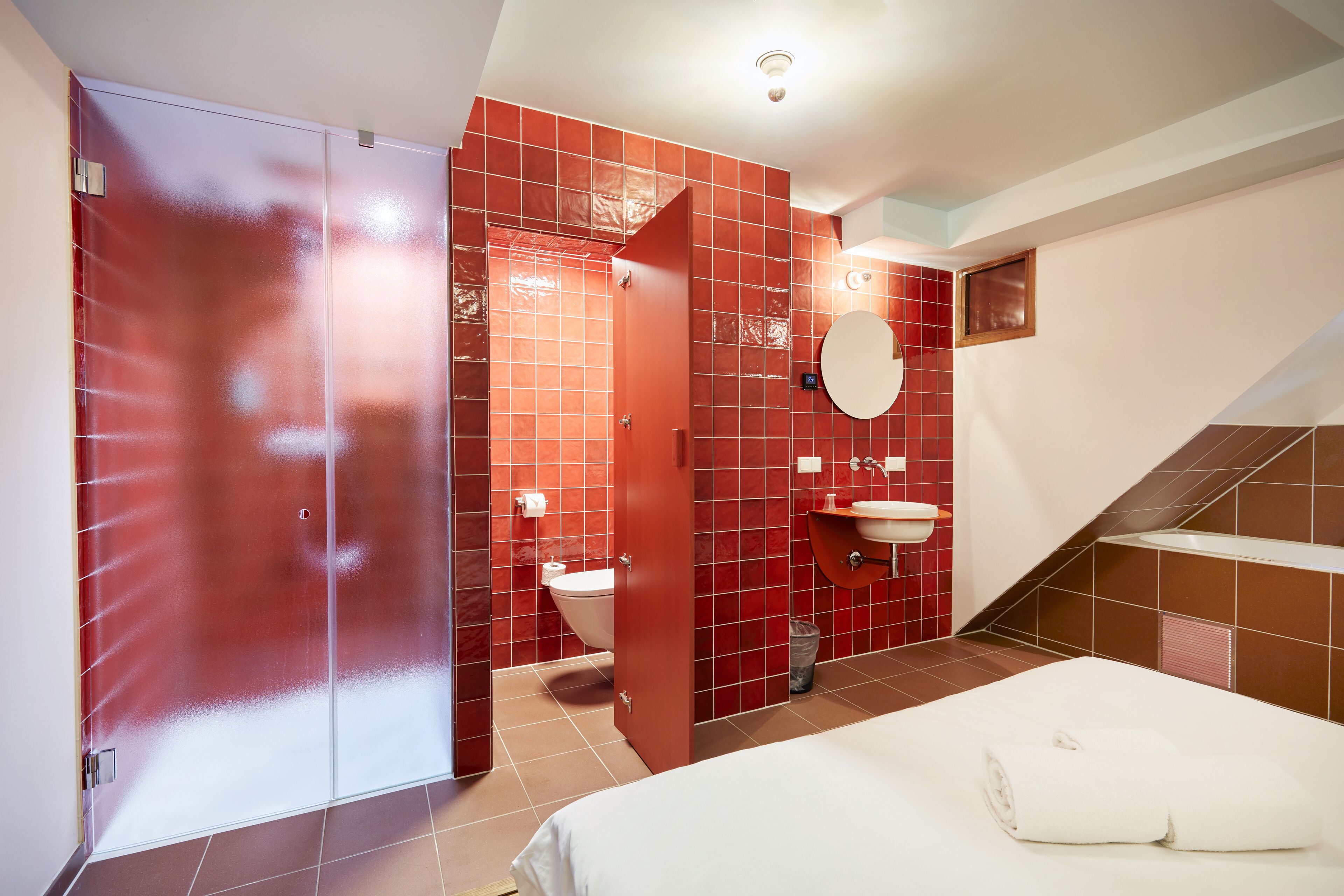 Quarto de hotel com cama, banheiro com vaso sanitário e pia, e parede com azulejos vermelhos.