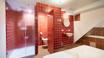 Quarto de hotel com cama, banheiro com vaso sanitário e pia, e parede com azulejos vermelhos.
