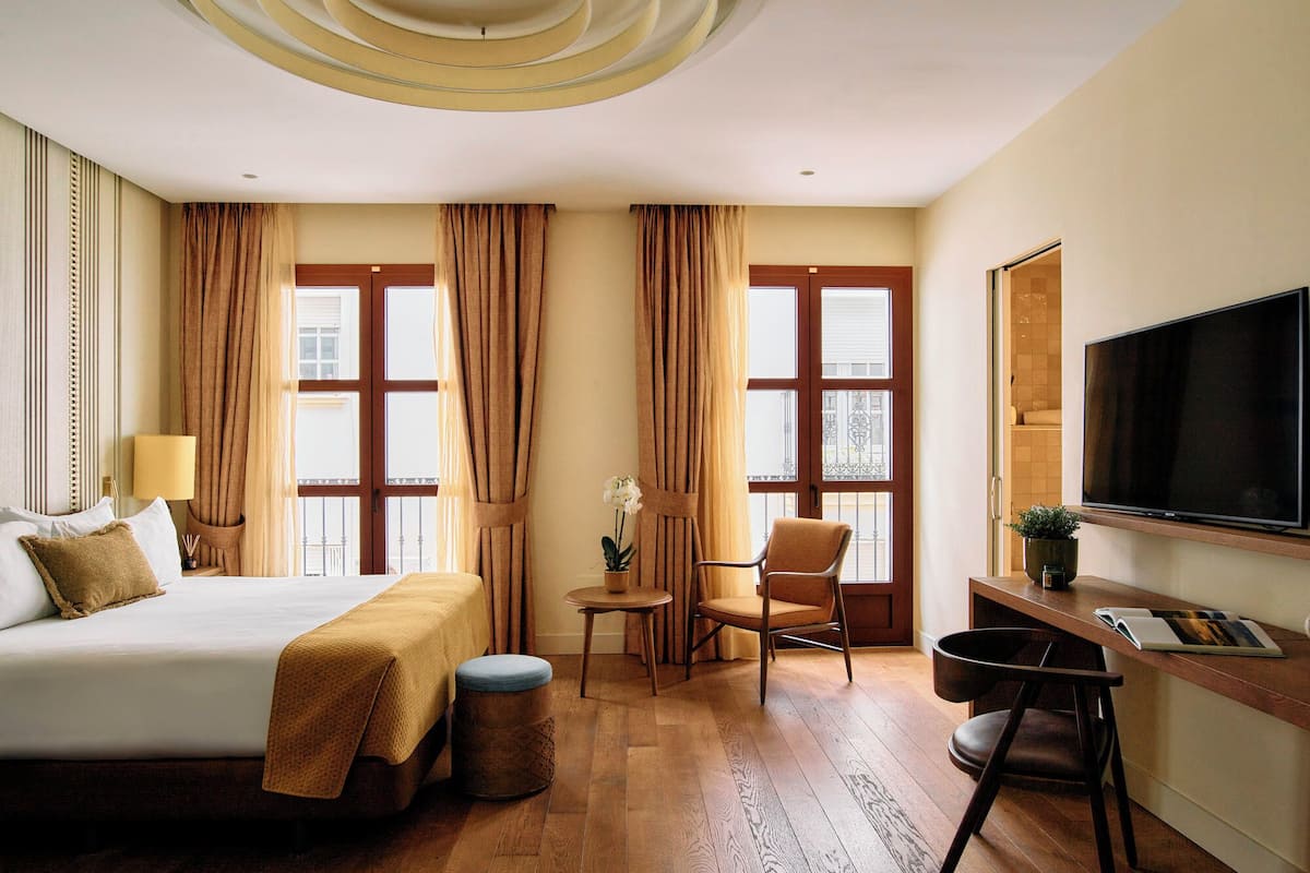 Quarto de hotel com uma cama grande, uma escrivaninha com televisão e uma cadeira. As janelas têm cortinas, e há uma pequena mesa com uma planta.