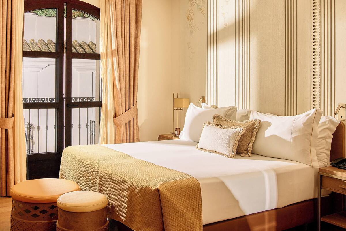 Quarto de hotel com cama, mesa de cabeceira, abajur e dois pufes.