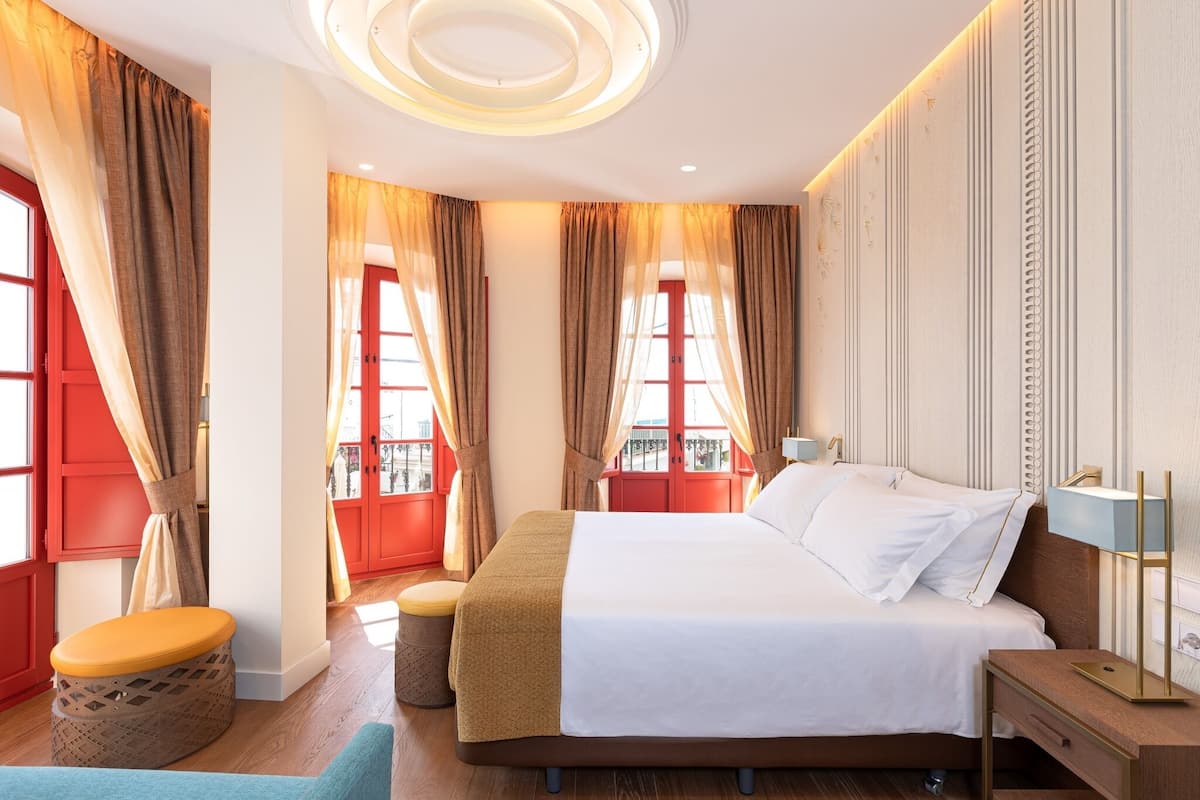Quarto de hotel com uma cama grande, mesa de cabeceira, luminária e vista da cidade através de grandes janelas.