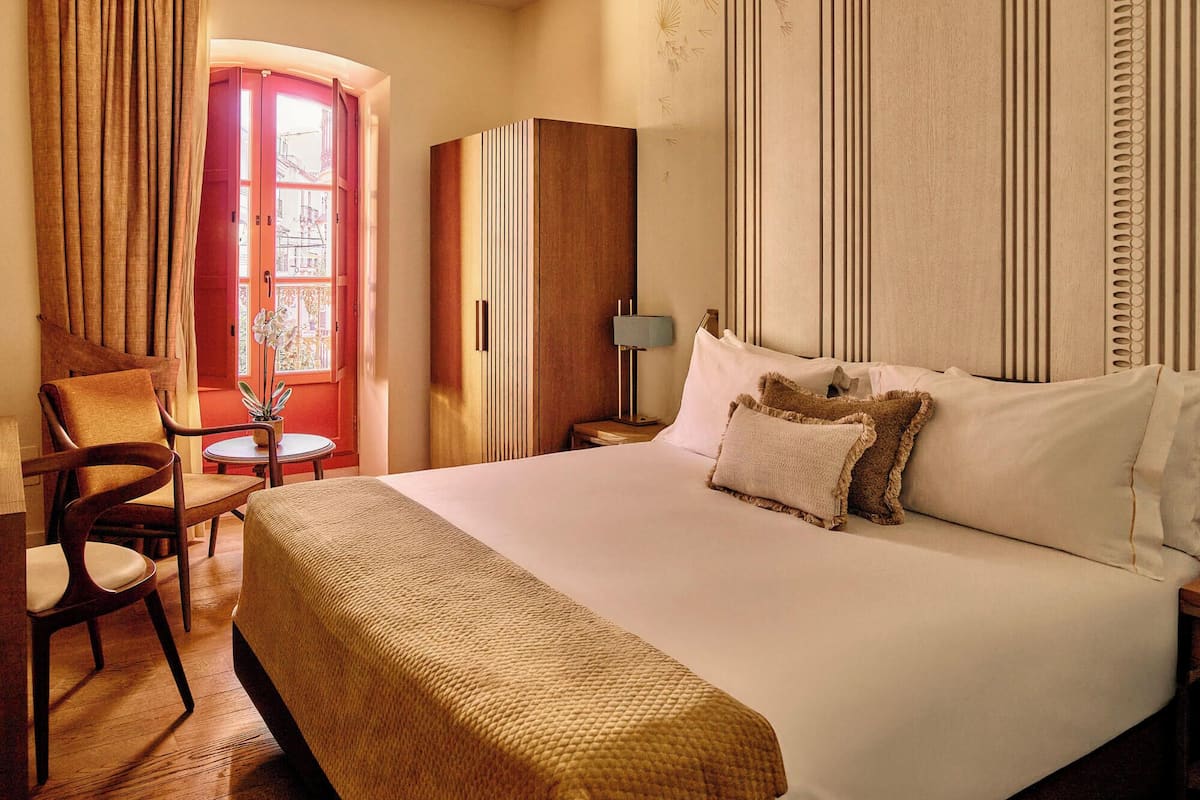 Quarto de hotel com uma cama grande, uma cadeira de madeira, uma mesinha e uma janela com cortinas.
