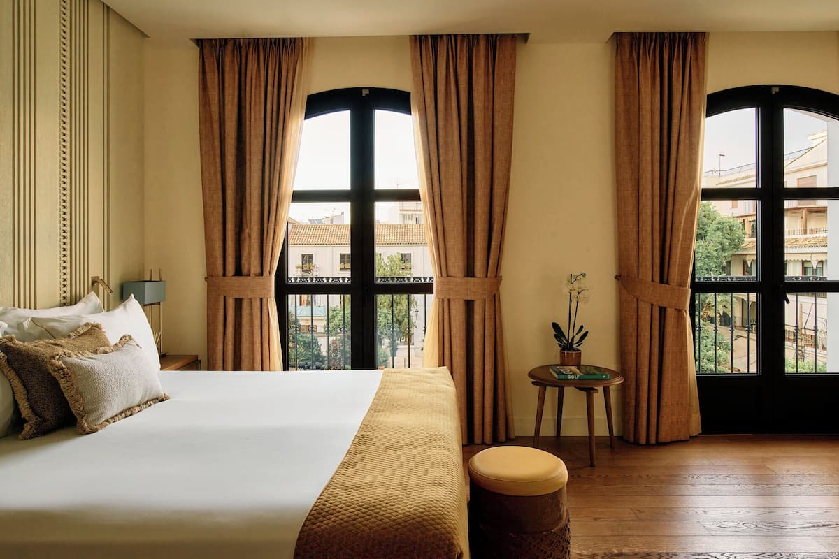 Quarto de hotel com uma cama grande, duas janelas com cortinas, uma mesinha com uma planta e vista para edifícios.