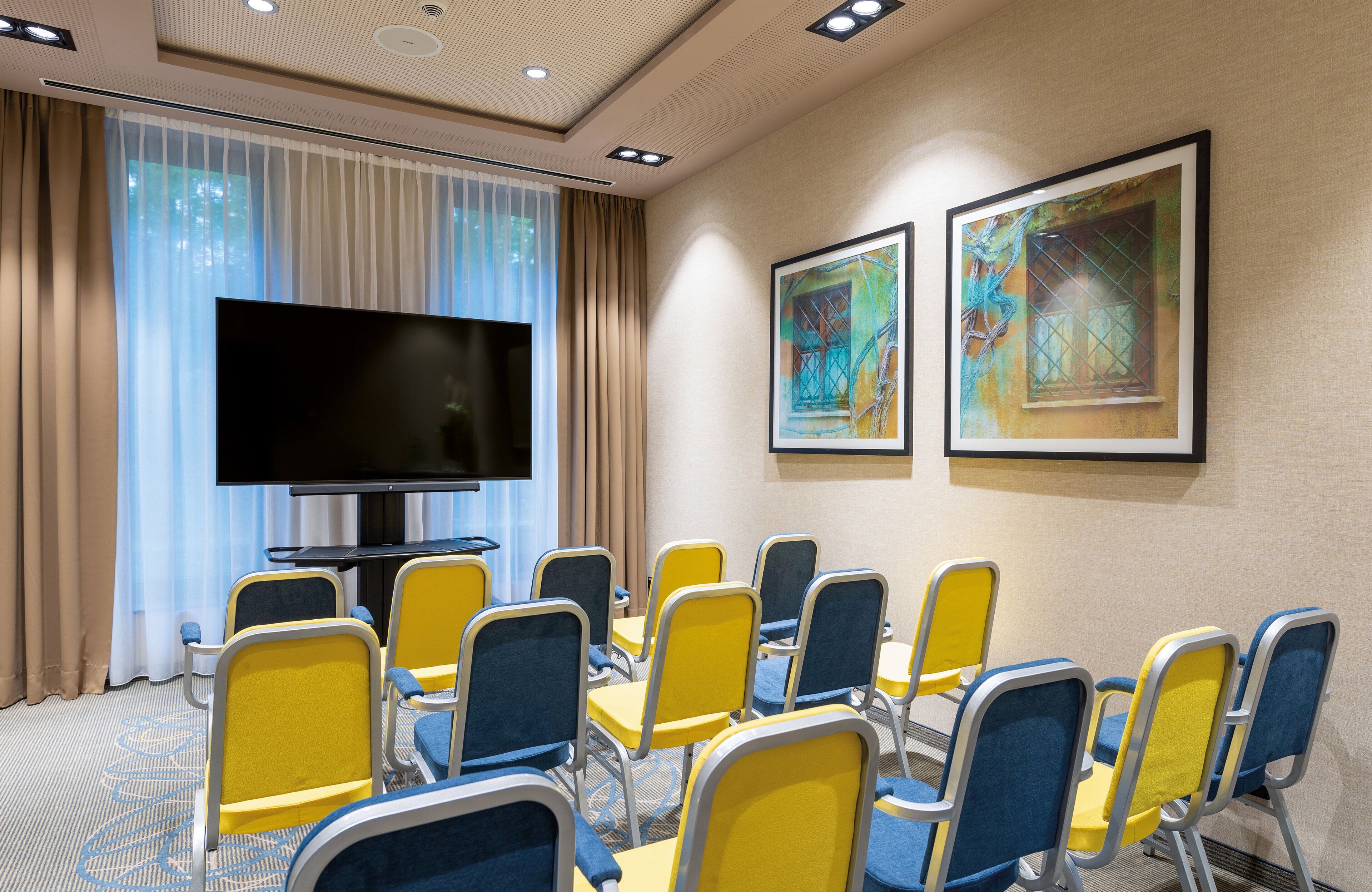 Uma sala de conferências com uma grande televisão de tela plana, cadeiras amarelas e azuis e duas pinturas abstratas emolduradas na parede.