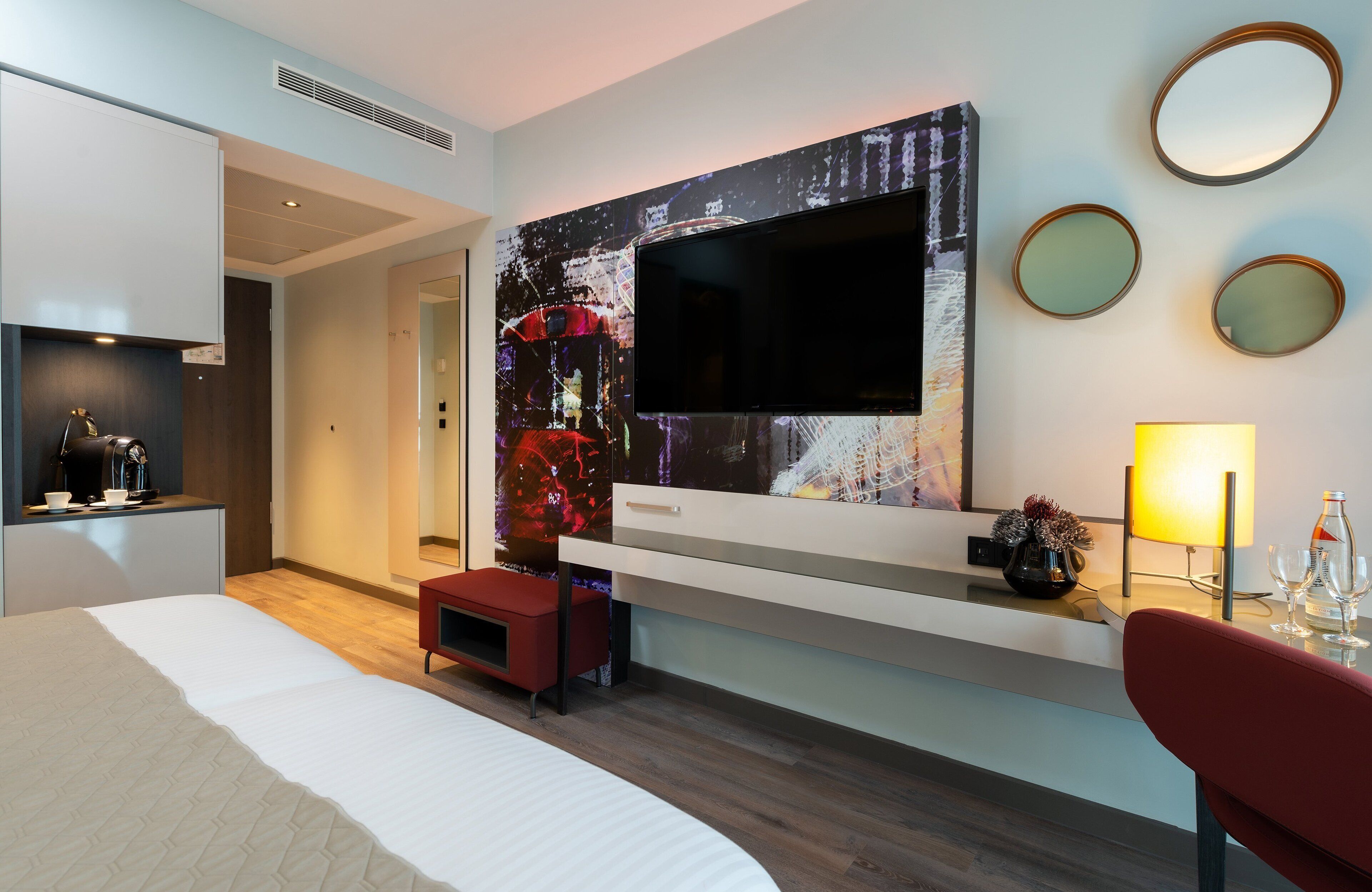 Quarto de hotel moderno com uma cama grande, televisão na parede, um pequeno espaço de estar e uma mesa com abajur.