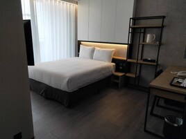 Quarto de hotel moderno com uma cama grande, mesas de cabeceira, uma escrivaninha com telefone e um armário com cafeteira.