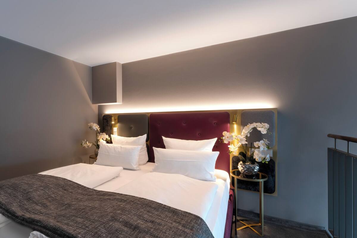 Quarto de hotel moderno com uma cama grande, cabeceira vermelha e uma mesinha de cabeceira com um vaso de flores.