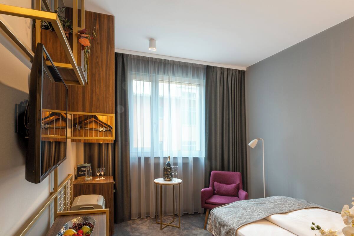 Quarto de hotel com uma cama, uma poltrona roxa, uma mesinha com uma garrafa e copos, uma televisão na parede e uma janela grande com cortinas.