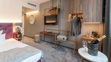 Quarto de hotel moderno com cama, escrivaninha, espelho, guarda-roupa e uma pequena mesa com champanhe e taças.