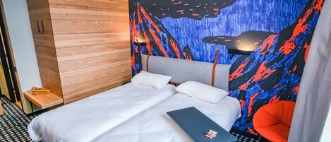 Quarto de hotel moderno com cama, laptop, guarda-roupa de madeira, um mural colorido e uma cadeira laranja pequena.