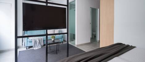 Quarto de hotel moderno com uma janela grande, uma cama, uma mesa redonda pequena com uma planta e um sofá azul ao fundo.