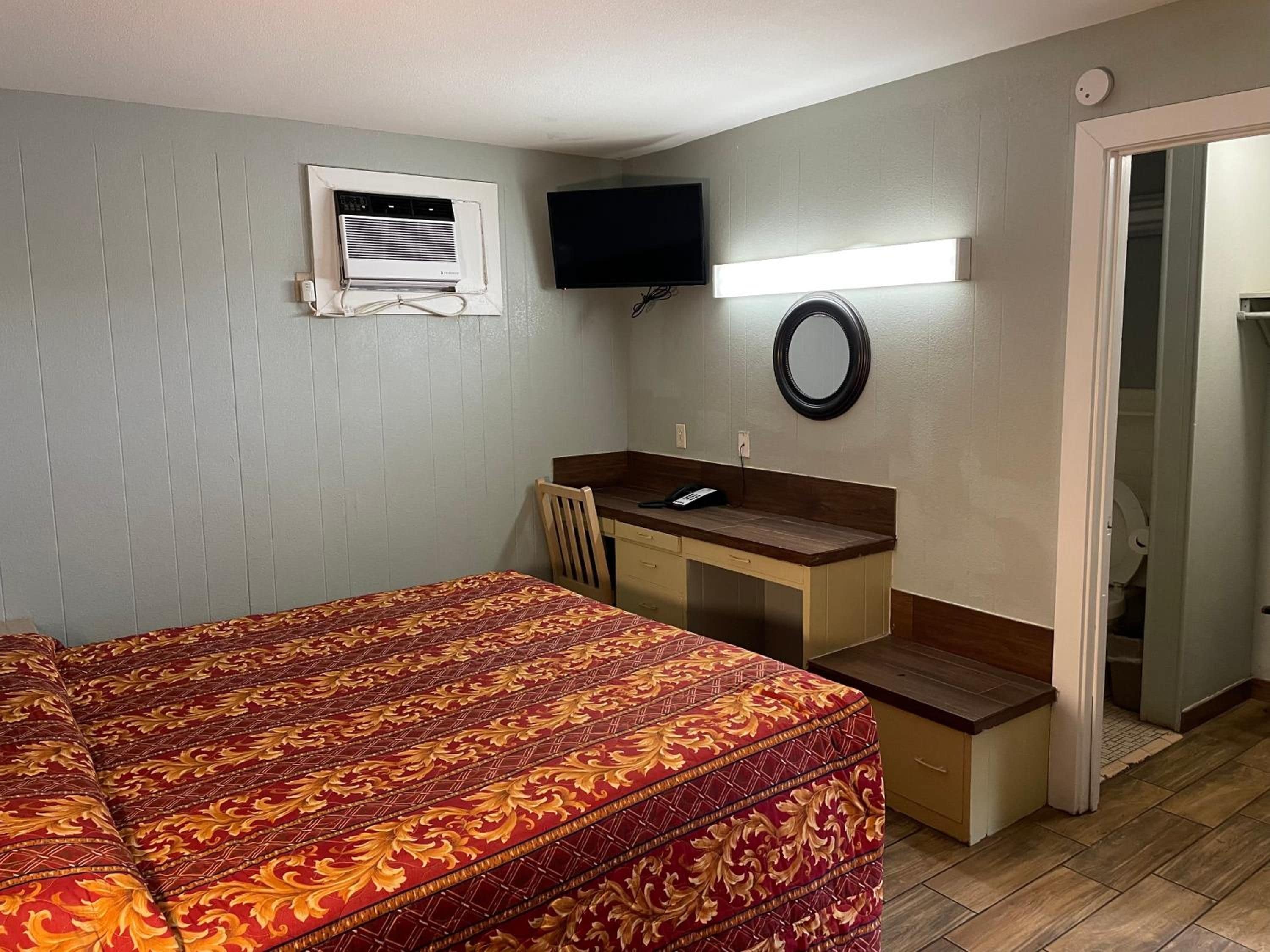 Quarto de hotel com cama, escrivaninha, cadeira, televisão, espelho e porta do banheiro.
