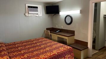 Quarto de hotel com cama, escrivaninha, cadeira, televisão, espelho e porta do banheiro.