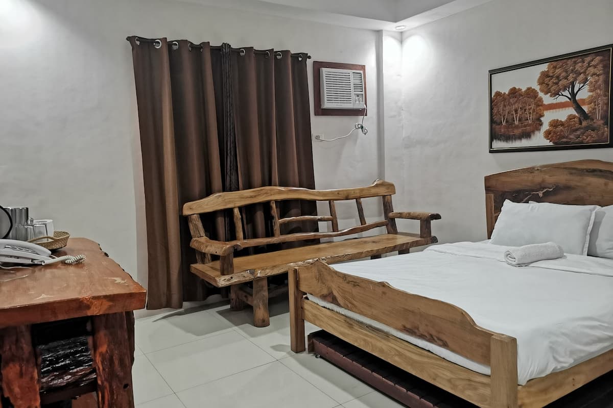 Quarto com cama de madeira, um banco de madeira, uma mesa pequena com telefone e um quadro na parede.