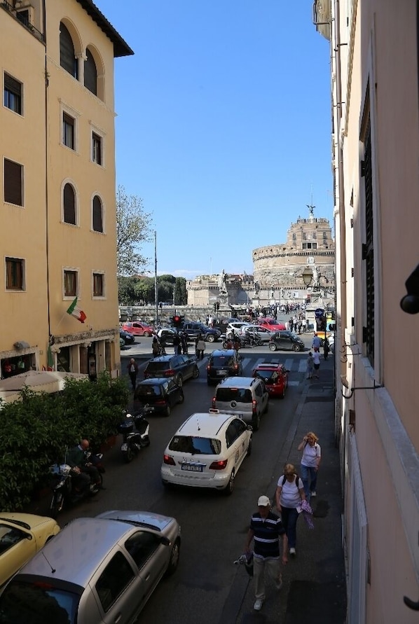Uma rua movimentada com carros e pedestres, edifícios com janelas em arco, e uma estrutura histórica imponente ao fundo.