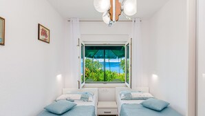 Quarto com duas camas, janela com vista para área verde e mar, e ventilador de teto.