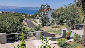 Uma área residencial com uma rua que leva a uma casa branca com telhado vermelho, cercada por vegetação e vista para o mar.
