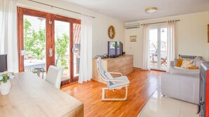 Uma sala de estar ampla, com piso de madeira, mesa de jantar, sofá, televisão e janela com cortinas.