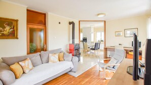 Uma sala de estar com sofá, cadeiras, TV e cozinha ao fundo.