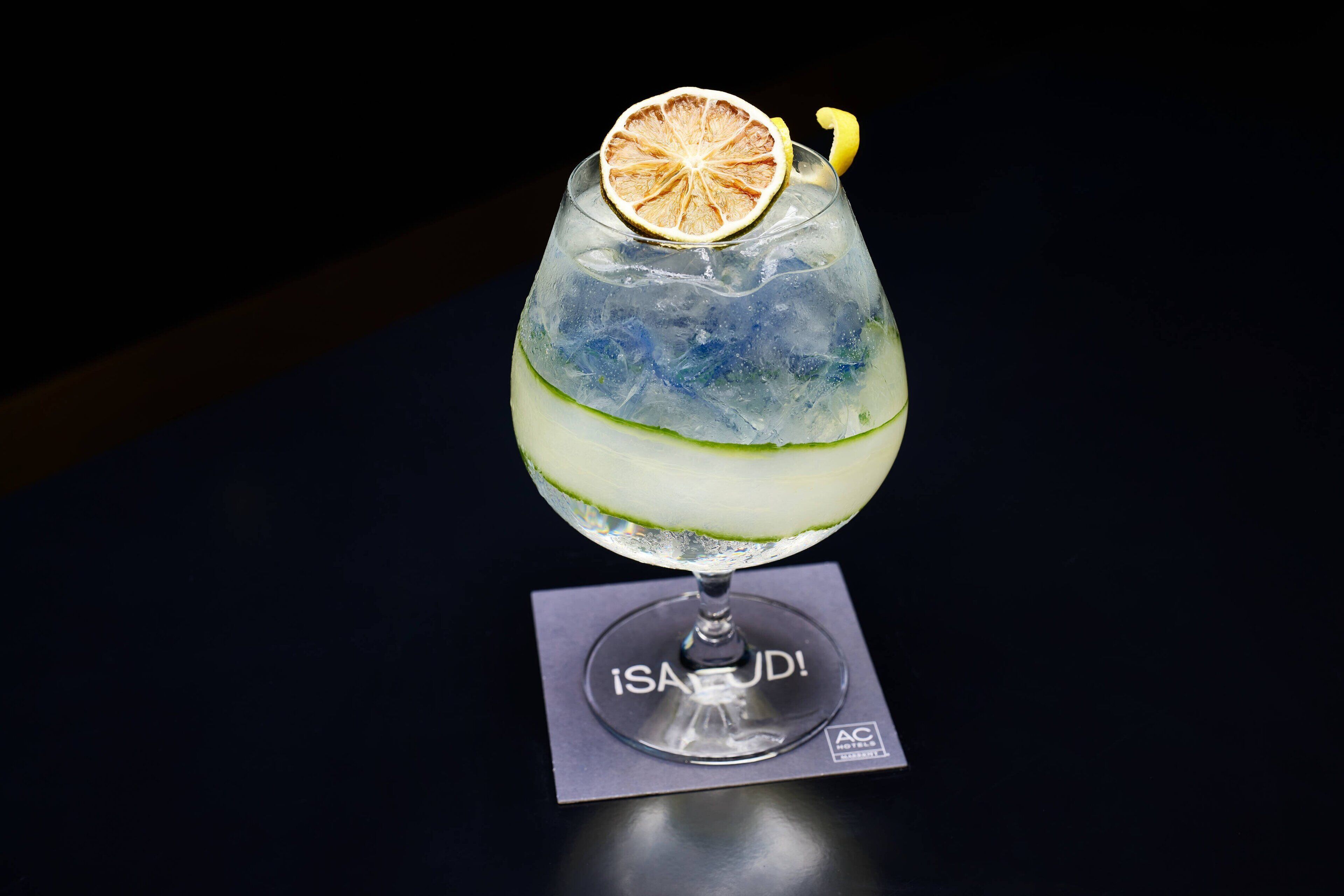 Um cocktail num copo com gelo, adornado com uma fatia de fruta cítrica e um twist de limão, sobre uma mesa preta com um cartão que diz "ISADU!".