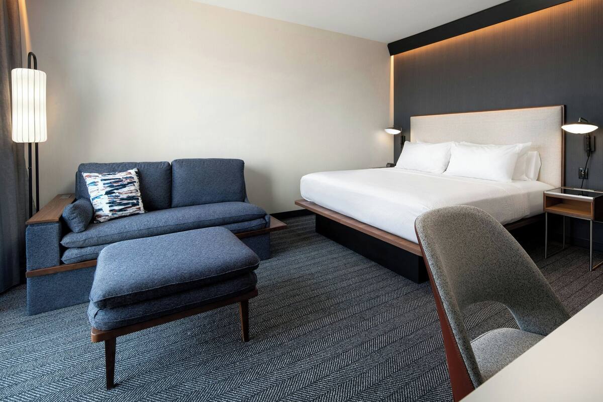 Quarto de hotel com cama, sofá, puf, cadeira, mesa de cabeceira e abajur.