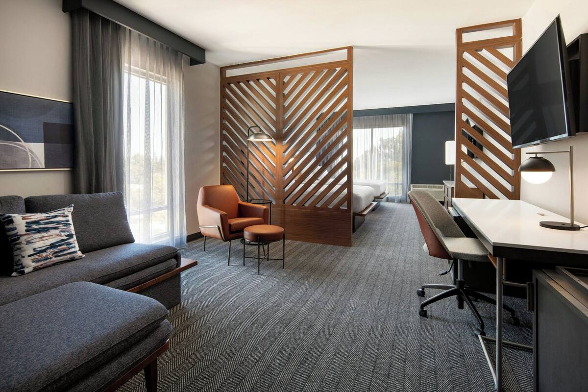 Quarto de hotel moderno com sofá, poltrona, escrivaninha e uma televisão grande.