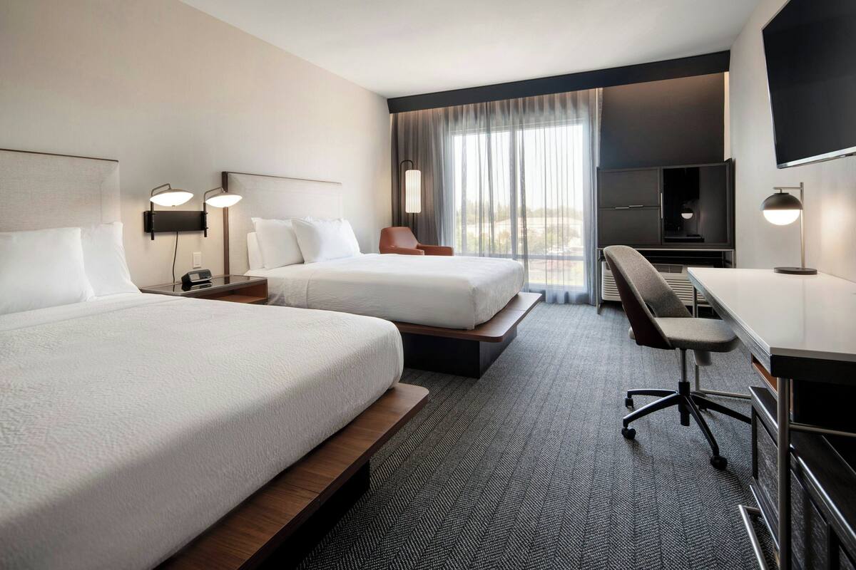 Quarto de hotel com duas camas, uma escrivaninha, uma cadeira, televisão e uma janela grande com cortinas.