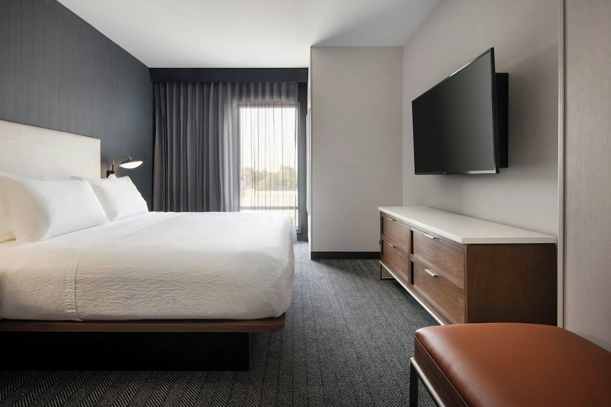 Quarto de hotel com uma cama grande, uma televisão de tela plana na parede, uma cômoda de madeira e uma janela com cortinas.