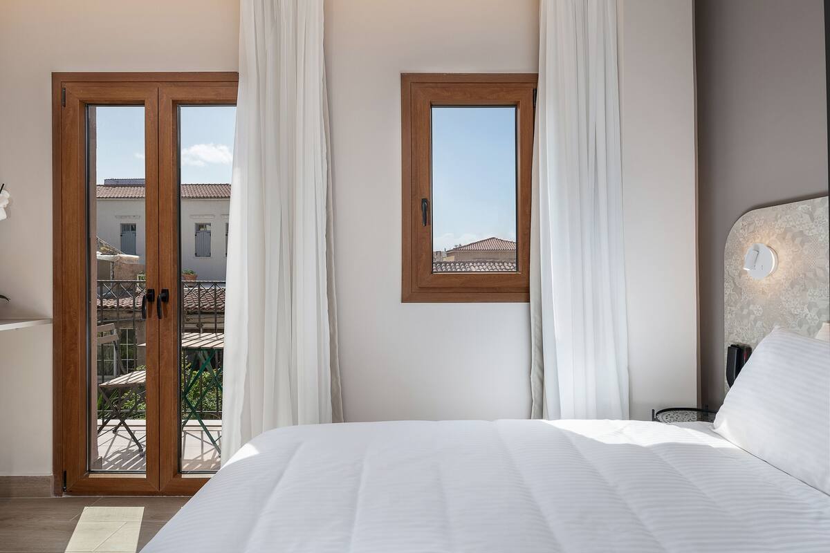 Quarto com uma cama grande, porta de madeira com painéis de vidro, uma janela pequena e vista para edifícios.