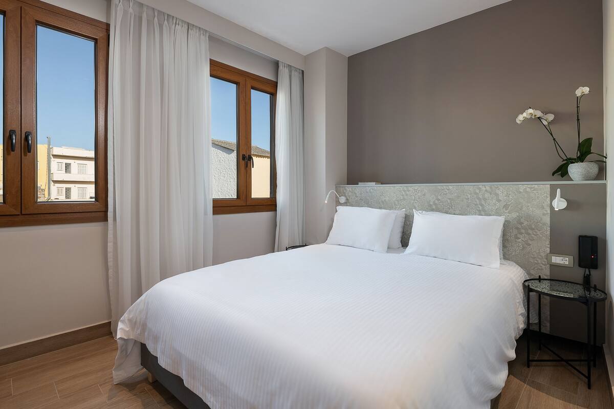 Quarto com cama grande, mesa de cabeceira, janela com cortinas e vista para prédios.