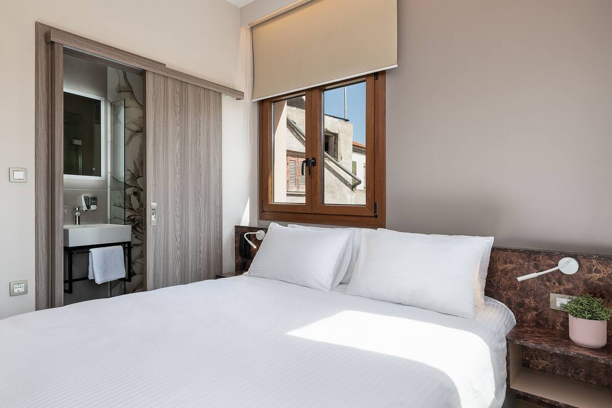 Quarto de hotel com cama, janela vista para edifícios, banheiro com pia e espelho, e mesa de cabeceira com uma planta.