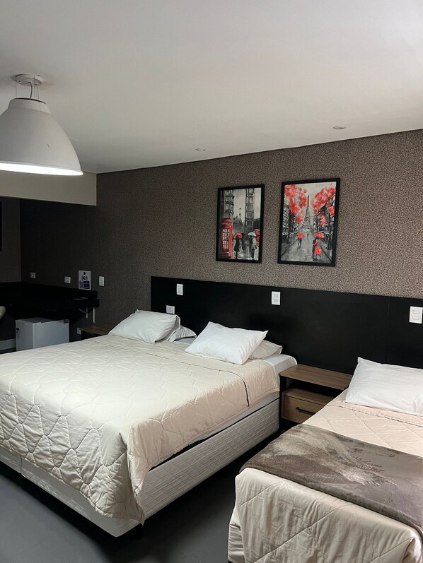 Quarto de hotel com uma cama grande, uma cama menor, uma escrivaninha e uma cadeira. Há duas pinturas na parede.