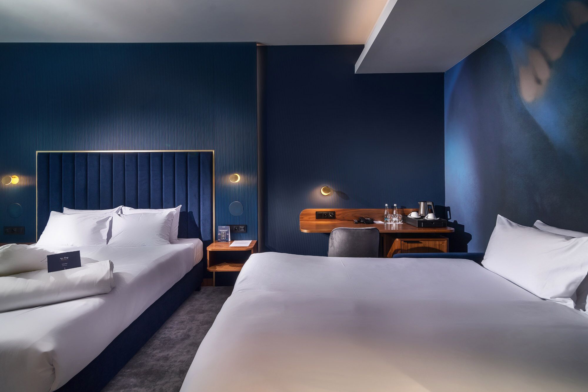 Quarto de hotel com duas camas, uma escrivaninha de madeira e televisão fixada na parede.