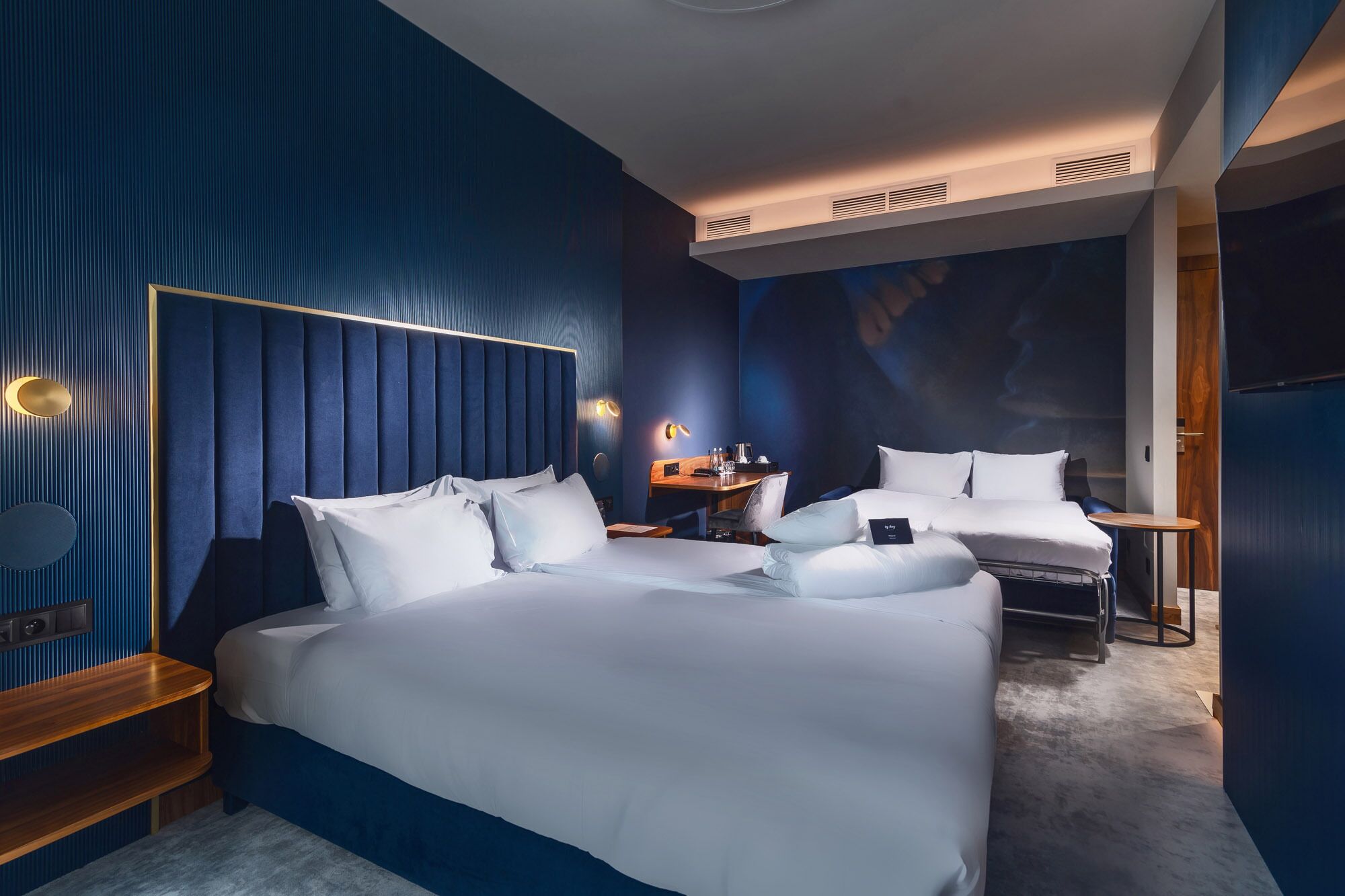 Quarto de hotel moderno com uma cama grande, uma cama menor, uma escrivaninha e um espaço de estar.