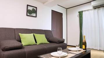 Sala de estar com sofá marrom, dois almofadas verdes, planta emoldurada, porta, janela com cortinas brancas e mesa com garrafa de vinho e copo.