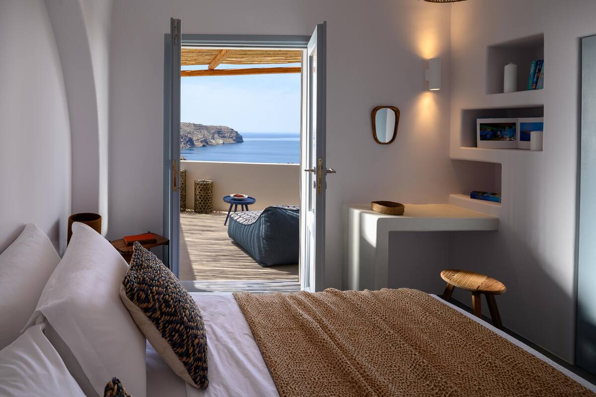 Quarto com cama grande, vista para o mar, pequeno espaço para sentar e banheiro com prateleira.