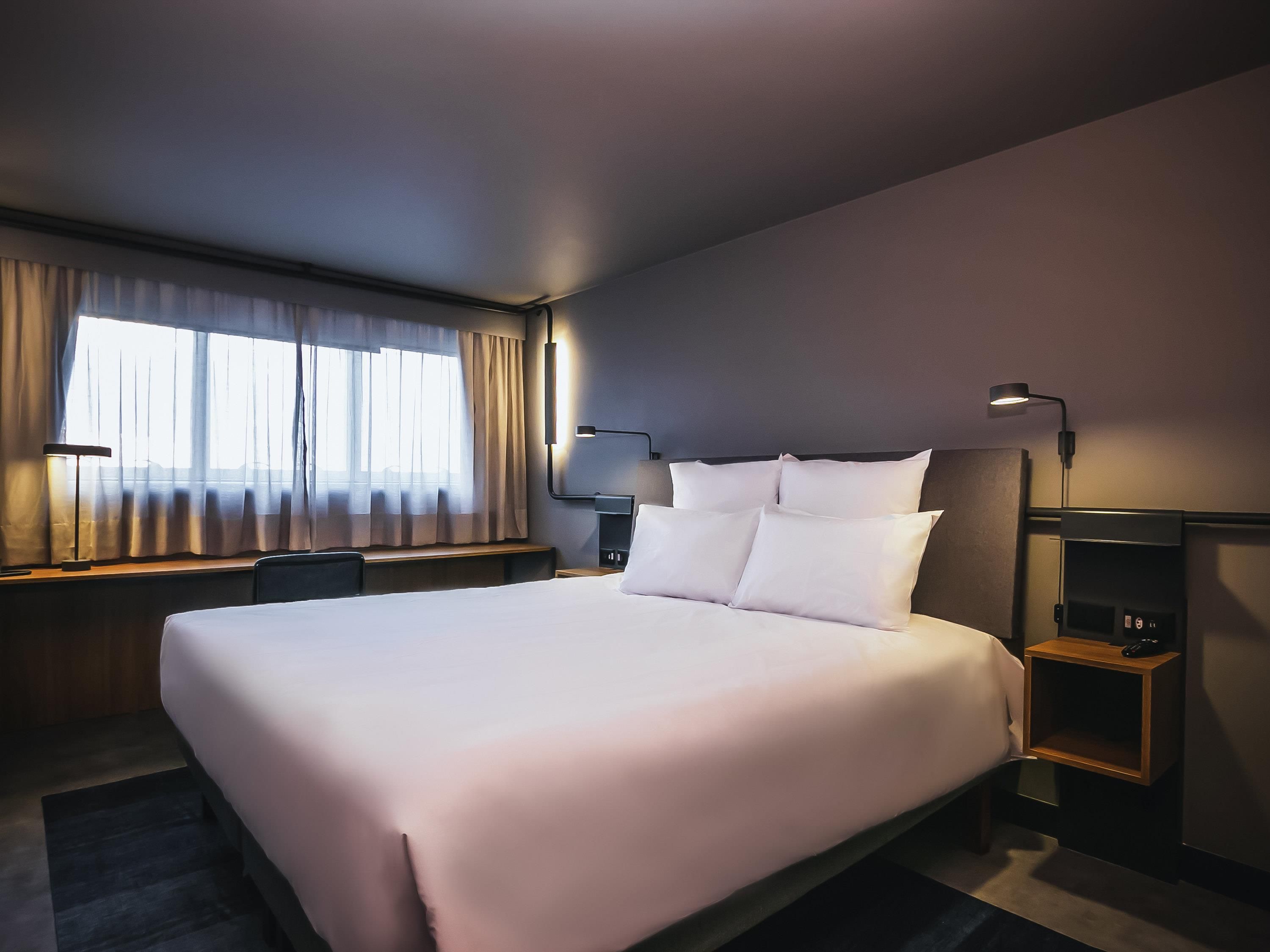Quarto de hotel com cama grande, mesas de cabeceira, escrivaninha, cadeira e janela com cortinas.