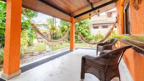 Um alpendre coberto ao ar livre com móveis de vime, uma rede e vista para um jardim e edifícios ao fundo.