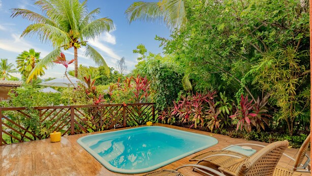 Uma área de piscina com deck de madeira, cadeiras de lounge de vime e vegetação tropical exuberante.