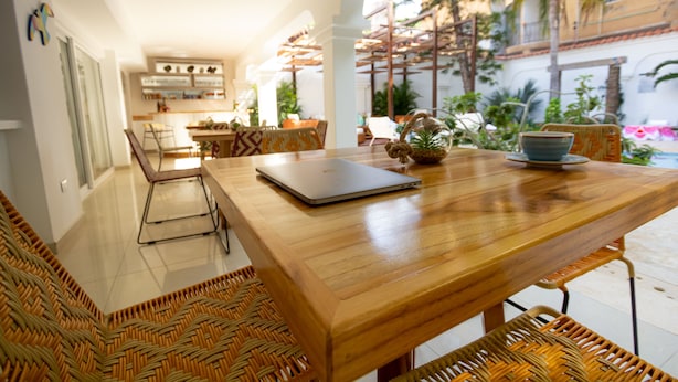 Uma mesa de jantar de madeira com um laptop, cadeiras de vime e vista para um pátio com plantas e uma varanda.