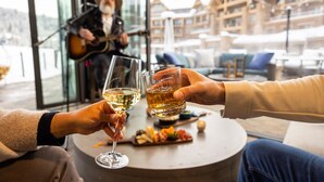 Duas pessoas brindando com bebidas à mesa, acompanhadas de uma tábua de queijos, em um quarto com grandes janelas que dão para um chalé.